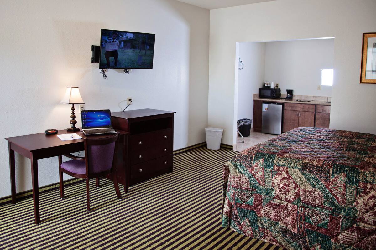 Quarto de hotel com cama, escrivaninha com um laptop, televisão, pequeno espaço de cozinha e lixeira.