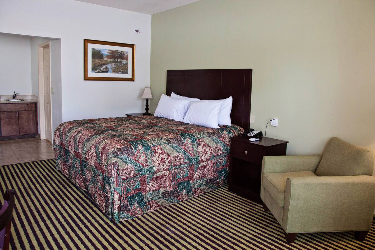 Quarto de hotel com uma cama grande, uma mesinha de cabeceira com abajur, uma cadeira, um quadro na parede e um banheiro visível por uma porta aberta.