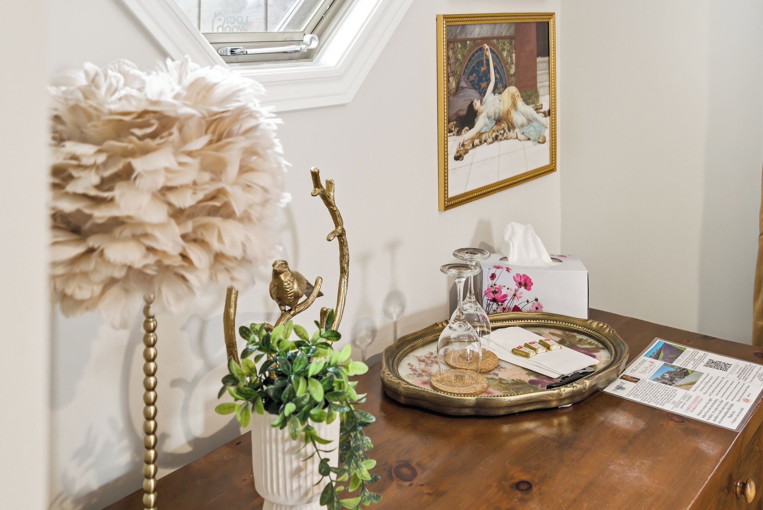 Uma mesa de madeira com uma bandeja decorativa, um vaso com folhagem verde, um objeto branco e fofo, um enfeite de pássaro dourado e um quadro na parede.