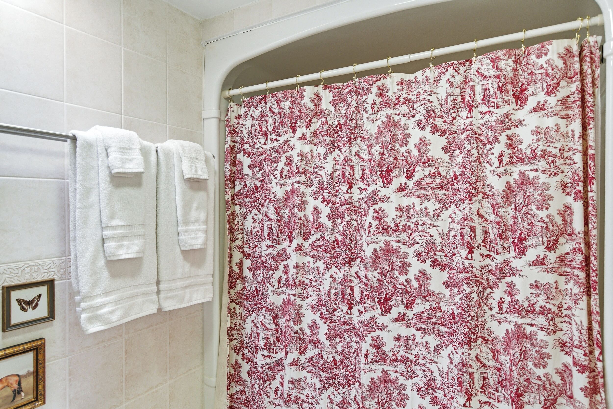 Um banheiro com cortina de chuveiro estampada em vermelho e branco, um porta-toalhas com toalhas brancas e quadros nas paredes.