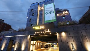 Um hotel moderno com um letreiro chamativo 'HOTEL SOL', um banner de 'GRANDE INAUGURAÇÃO' e um jardim no terraço.