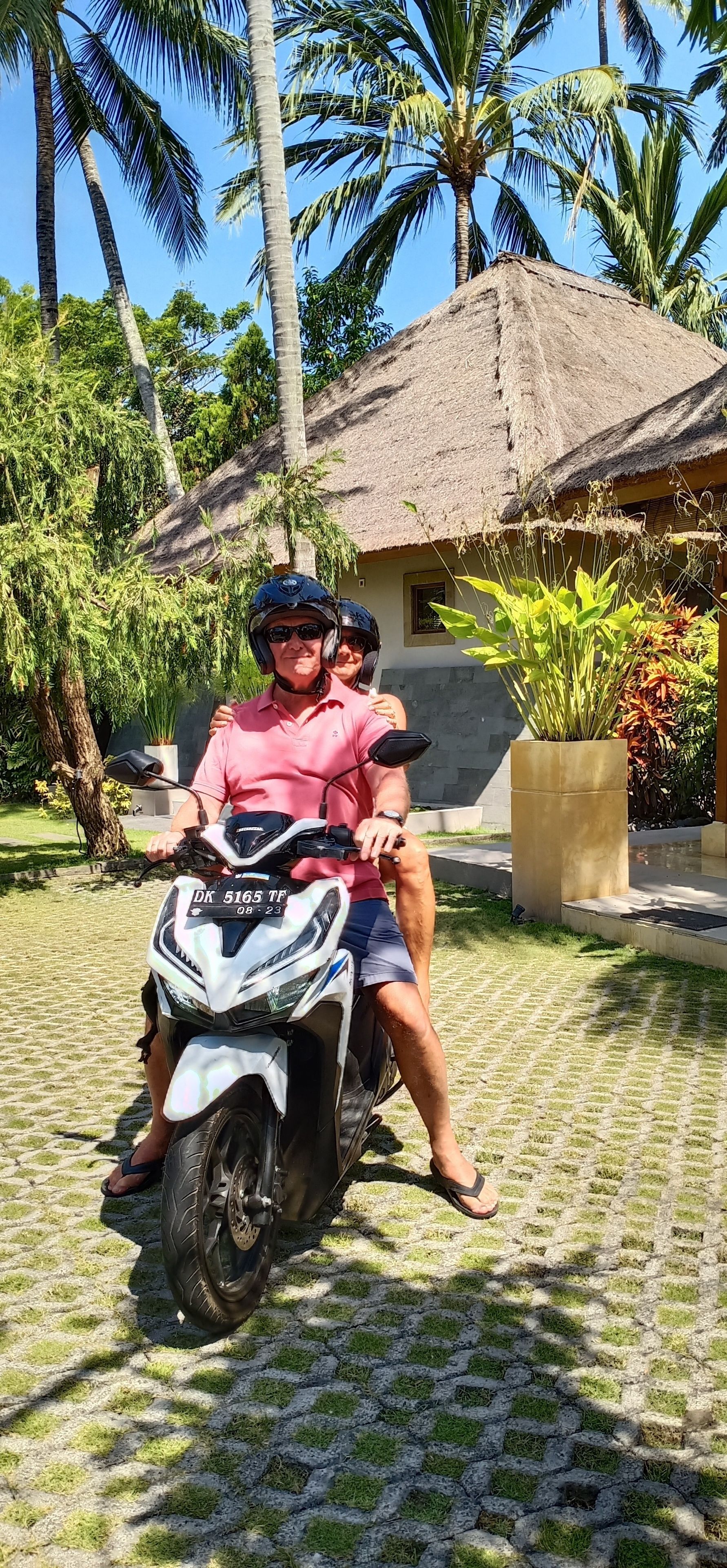 Uma pessoa conduzindo um scooter com placa DK 5165 TF, estacionado em um caminho de paralelepípedos com um edifício de telhado de palha e palmeiras ao fundo.
