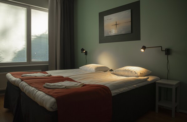 Uma cama bem arrumada com um cobertor e travesseiros vermelhos, duas luminárias de cabeceira, um criado-mudo branco e um quadro na parede.