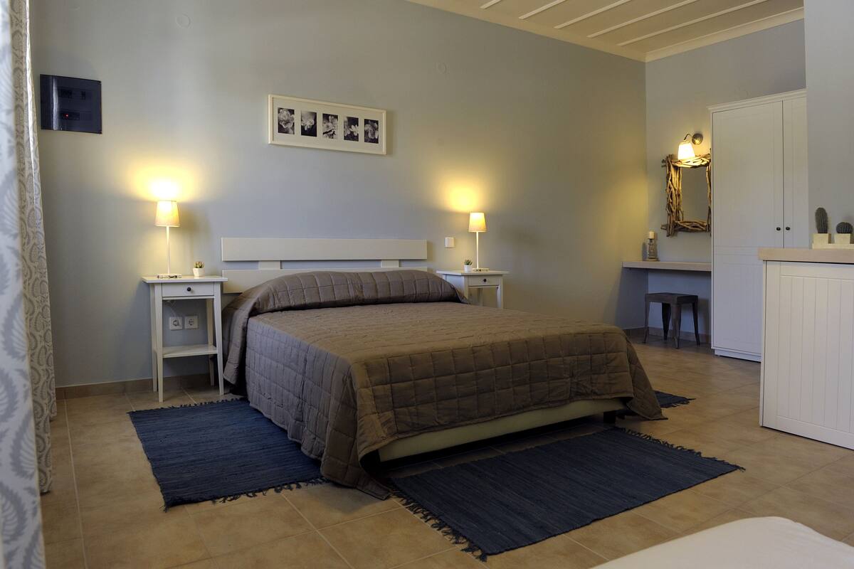 Quarto com cama, duas mesas de cabeceira com luminárias, uma cômoda, escrivaninha com cadeira, guarda-roupa e espelho fixado na parede.