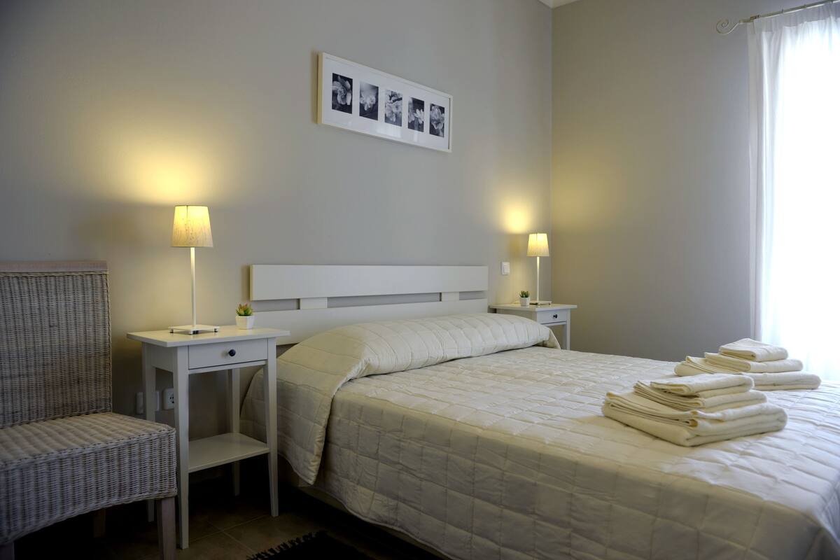 Cama bem arrumada com colcha branca, duas mesas de cabeceira com abajures, uma cadeira e uma fotografia emoldurada na parede.