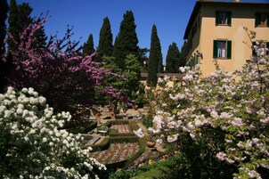 Um jardim com sebes bem aparadas e plantas floridas, um banco branco e um edifício com persianas verdes.