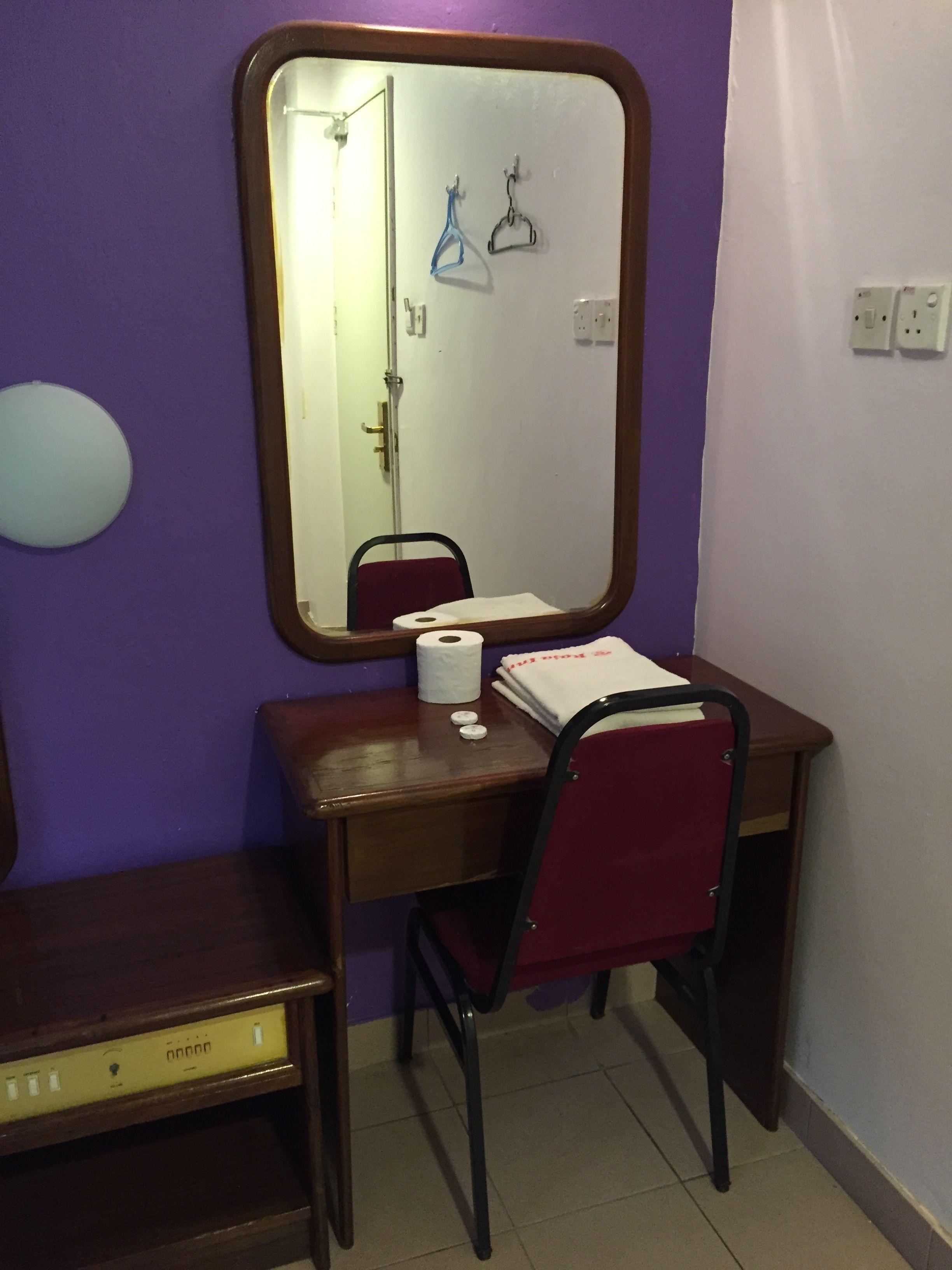 Quarto pequeno com uma escrivaninha, cadeira e espelho. Há um rolo de papel higiénico e uma pequena caixa na escrivaninha.