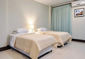 Quarto de hotel com duas camas, uma mesa de cabeceira, abajur, janela com cortinas e ar-condicionado fixado na parede.