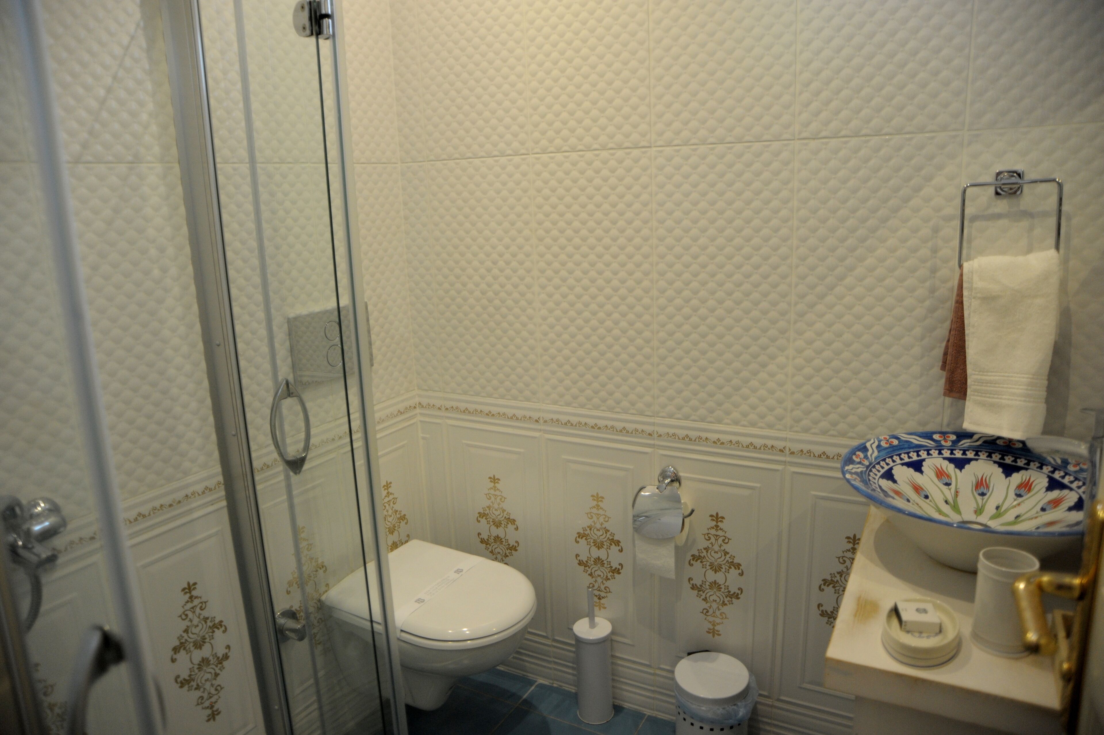 Casa de banho com cabine de duche em vidro, sanita, lavatório com bacia decorativa e um porta-toalhas.