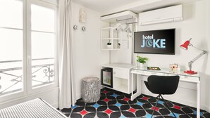 Quarto de hotel com uma janela grande, uma escrivaninha com um computador, uma cadeira, um pequeno armário e uma TV exibindo "hotel JOKE ASTOTEL PARIS".