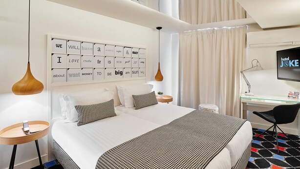Quarto de hotel moderno com cama bem arrumada, escrivaninha com cadeira e televisão.