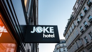 Placa do hotel JOKE, com design moderno, localizado em uma cidade com arquitetura clássica.