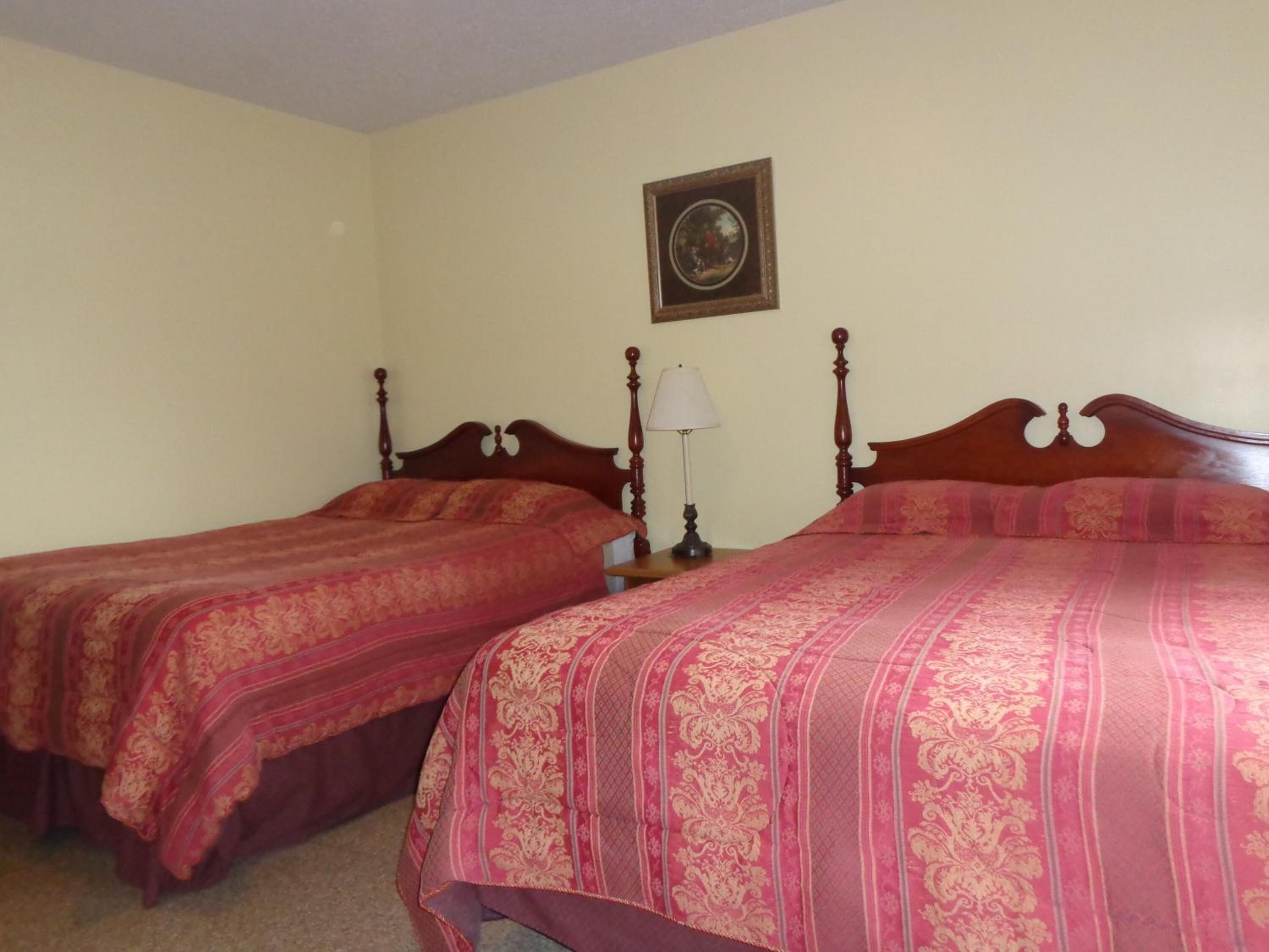 Duas camas de solteiro com cabeceiras de madeira e roupa de cama vermelha combinando em um quarto com paredes lisas e um quadro emoldurado sobre as camas.