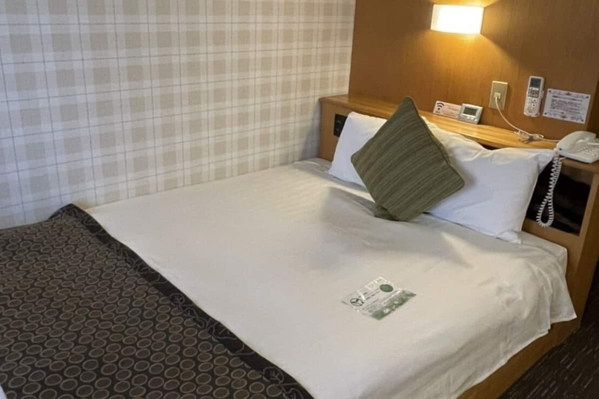 Quarto de hotel com cama, mesa de cabeceira, telefone e luminária fixada na parede.