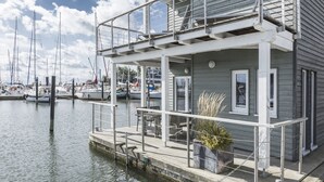Um edifício à beira-mar com varanda, um cais com barcos e uma pequena área de estar ao ar livre.