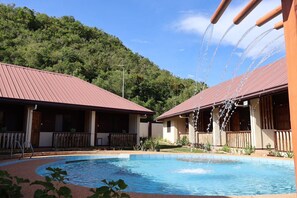 Uma área de piscina com dois edifícios, cada um com telhado vermelho e varandas de madeira, rodeada por vegetação e um cenário montanhoso.