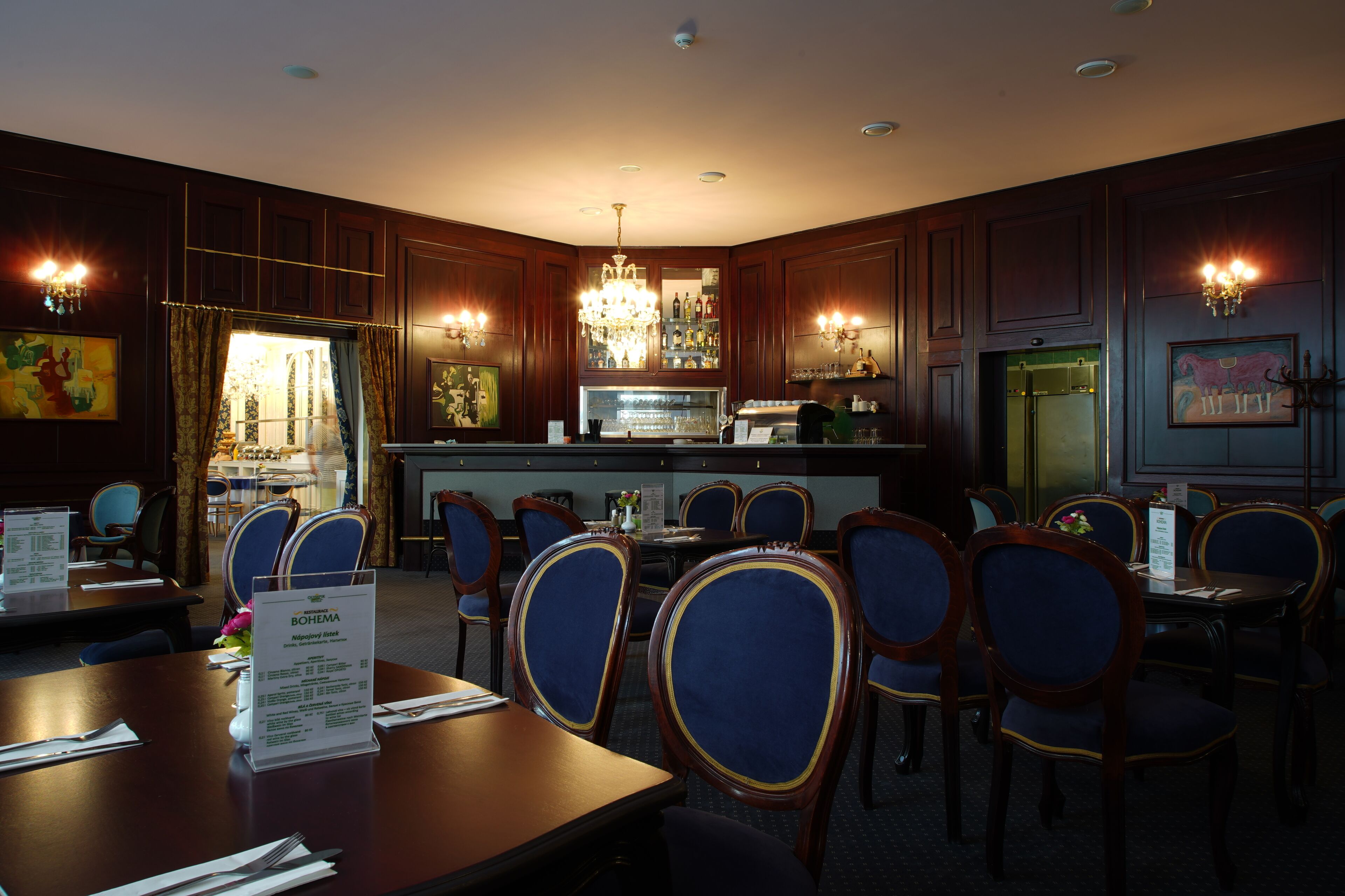 Um restaurante com painéis de madeira, cadeiras estofadas em azul, área de bar e um candelabro.
