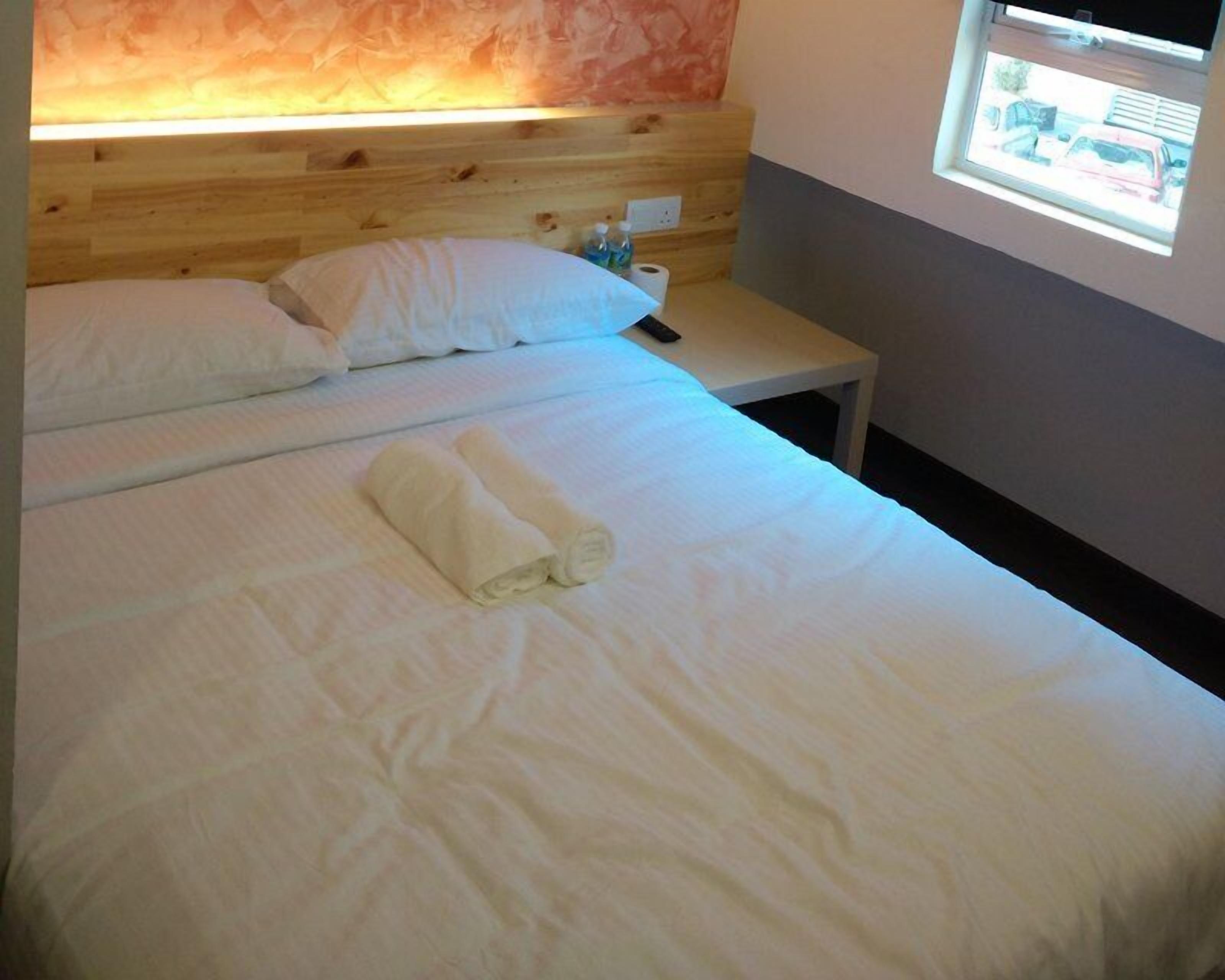 Uma cama bem arrumada com roupa de cama branca e uma toalha enrolada, uma mesa de cabeceira com um controle remoto e uma janela com vista para carros estacionados.