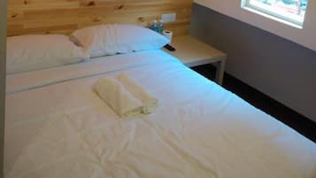Uma cama bem arrumada com roupa de cama branca e uma toalha enrolada, uma mesa de cabeceira com um controle remoto e uma janela com vista para carros estacionados.
