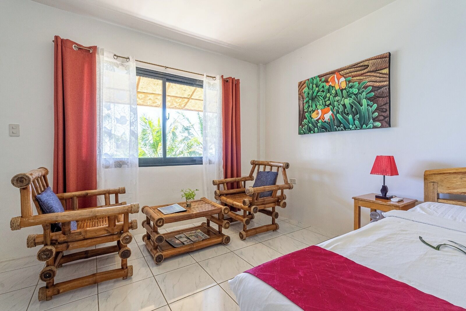 Quarto com cama, duas cadeiras de madeira, uma mesinha, mesa de cabeceira com abajur e janela com cortinas.