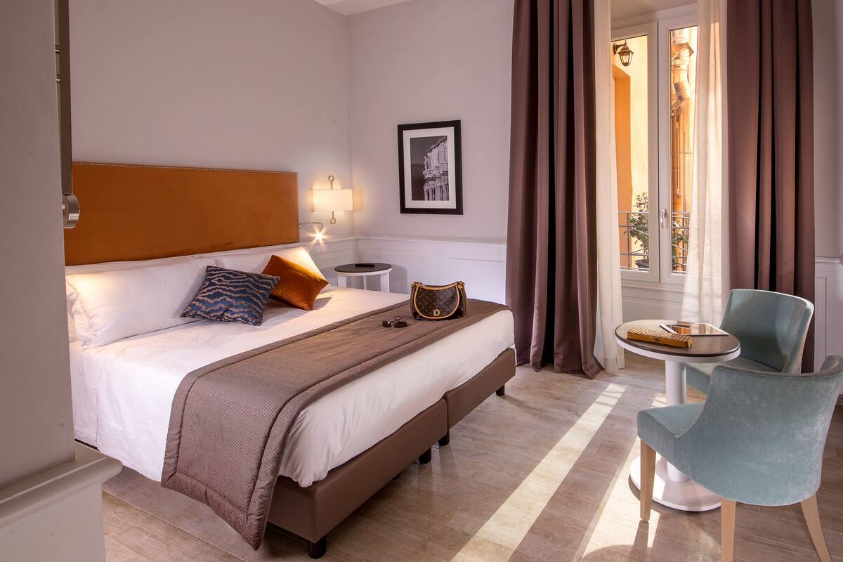 Quarto de hotel com cama, mesa de cabeceira, luminária, cadeira e vista para o exterior.