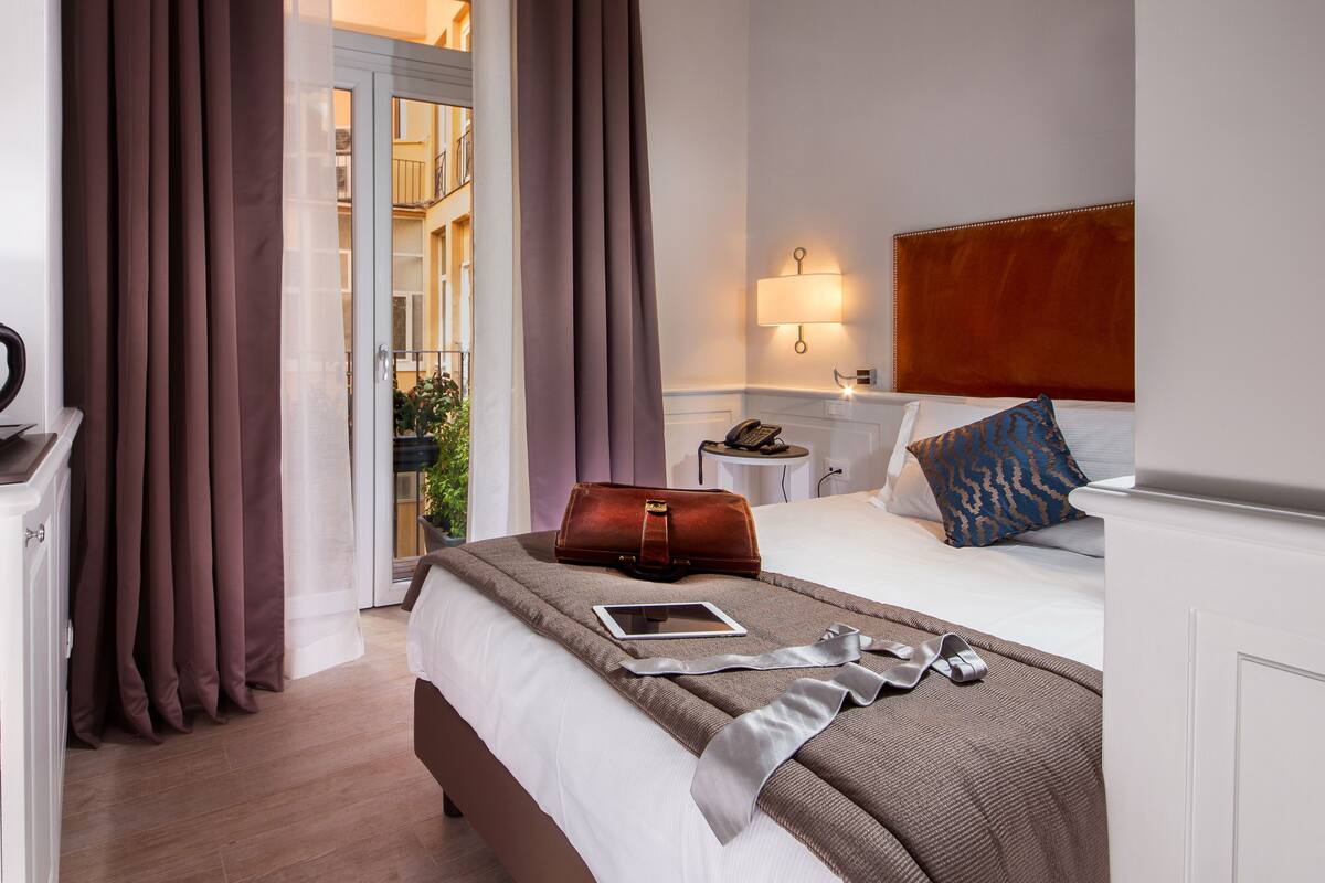Quarto de hotel com uma cama, uma mesa de cabeceira com abajur, um telefone e uma mala de couro marrom sobre a cama.