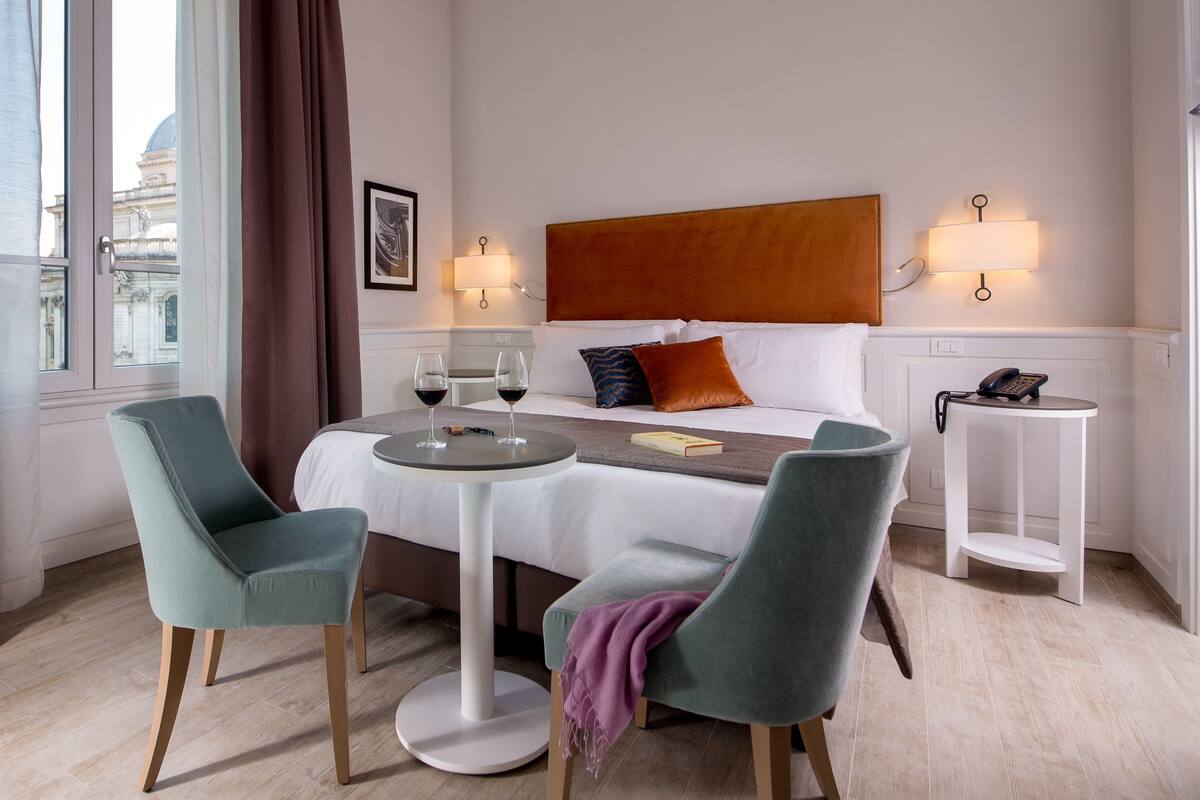 Quarto de hotel com uma cama, duas cadeiras, uma mesinha, uma mesinha de cabeceira com telefone e vista para um prédio pela janela.