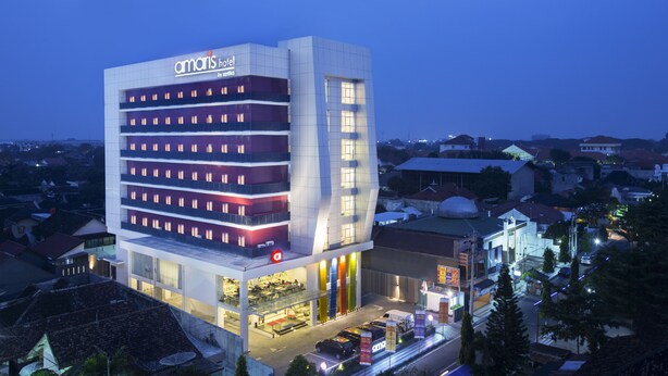 Um hotel de vários andares com design moderno, exibindo um letreiro 'Amaris Hotel' bem visível no topo. O edifício possui várias varandas e um restaurante no nível térreo.