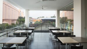 Um espaço de refeições interno com mesas e cadeiras, um ventilador de teto e vista para edifícios e vegetação lá fora.