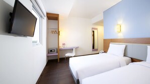 Quarto de hotel com duas camas, televisão fixada na parede, secretária com espelho e um espaço de casa de banho pequeno.