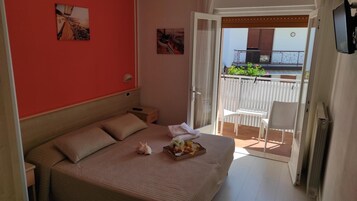 Quarto com cama, mesa de cabeceira e pequena mesa com bandeja de comida. Há uma varanda com cadeira e mesa.