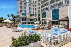 Área de piscina de hotel com espreguiçadeiras e guarda-sóis, cercada por um edifício com vários andares e varandas.
