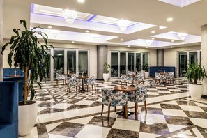Um saguão de hotel com piso xadrez, cadeiras florais e plantas em vasos.