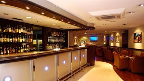 Uma área de bar com balcão polido, prateleiras com garrafas de bebidas e um espaço de estar com cadeiras e uma televisão.