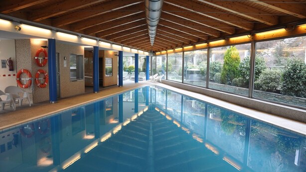 Piscina interior com água azul cristalina, vigas de madeira no teto e grandes janelas com vista para o verde lá fora.