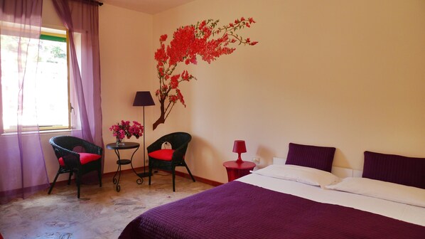 Quarto de hotel com uma cama, duas cadeiras, uma mesinha com um vaso de flores, uma luminária e um mural na parede.