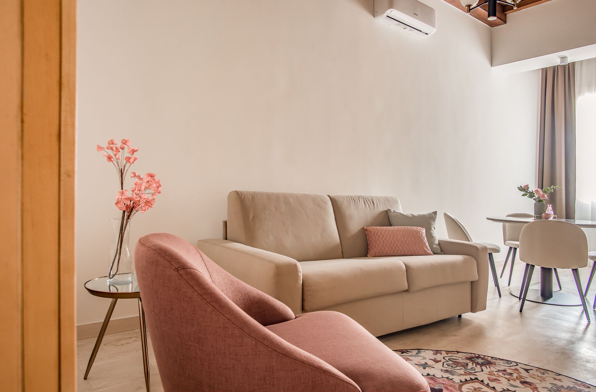 Uma sala de estar moderna com um sofá bege, uma poltrona rosa, uma pequena mesa de vidro com um vaso de flores rosa, e uma área de jantar com mesa redonda e cadeiras.