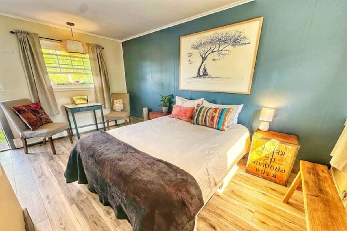 Um quarto com uma cama grande, um banquinho, uma mesa pequena, uma cadeira e uma janela com cortinas.