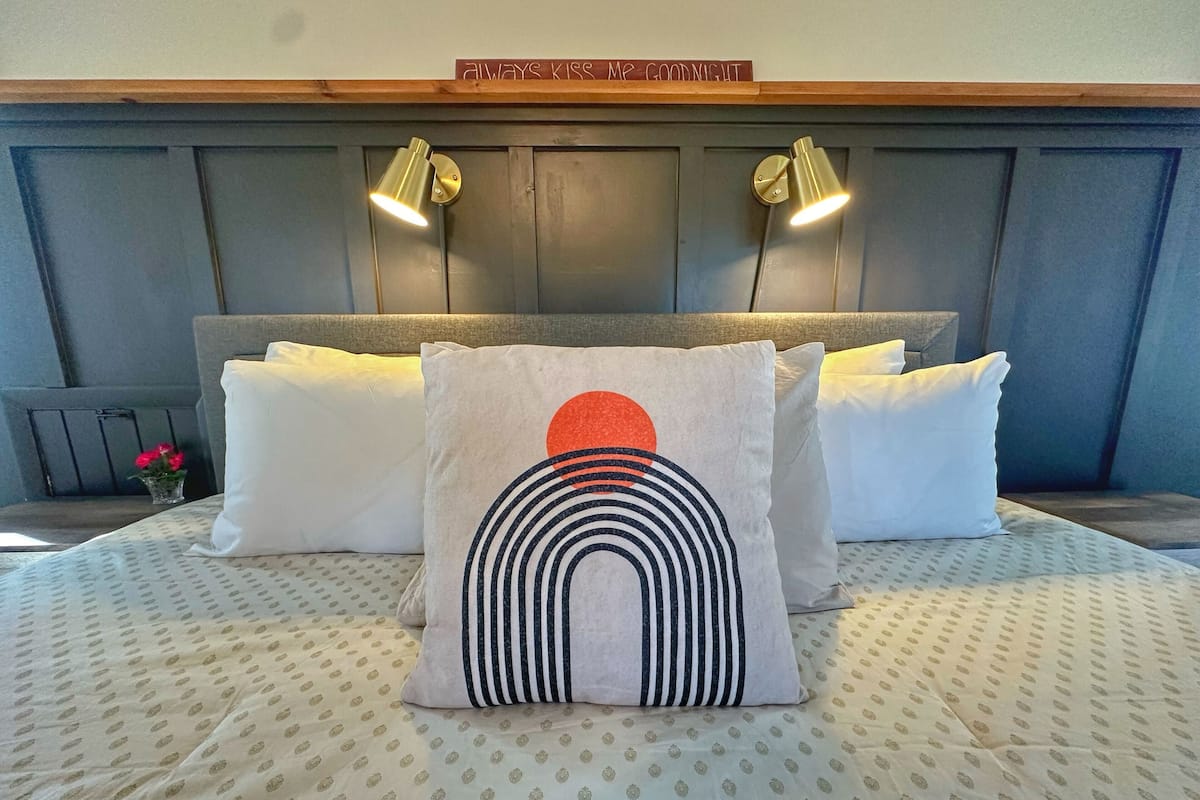 Cama bem arrumada com roupa de cama estampada, duas luminárias de cabeceira e um travesseiro decorativo com um desenho de sol.