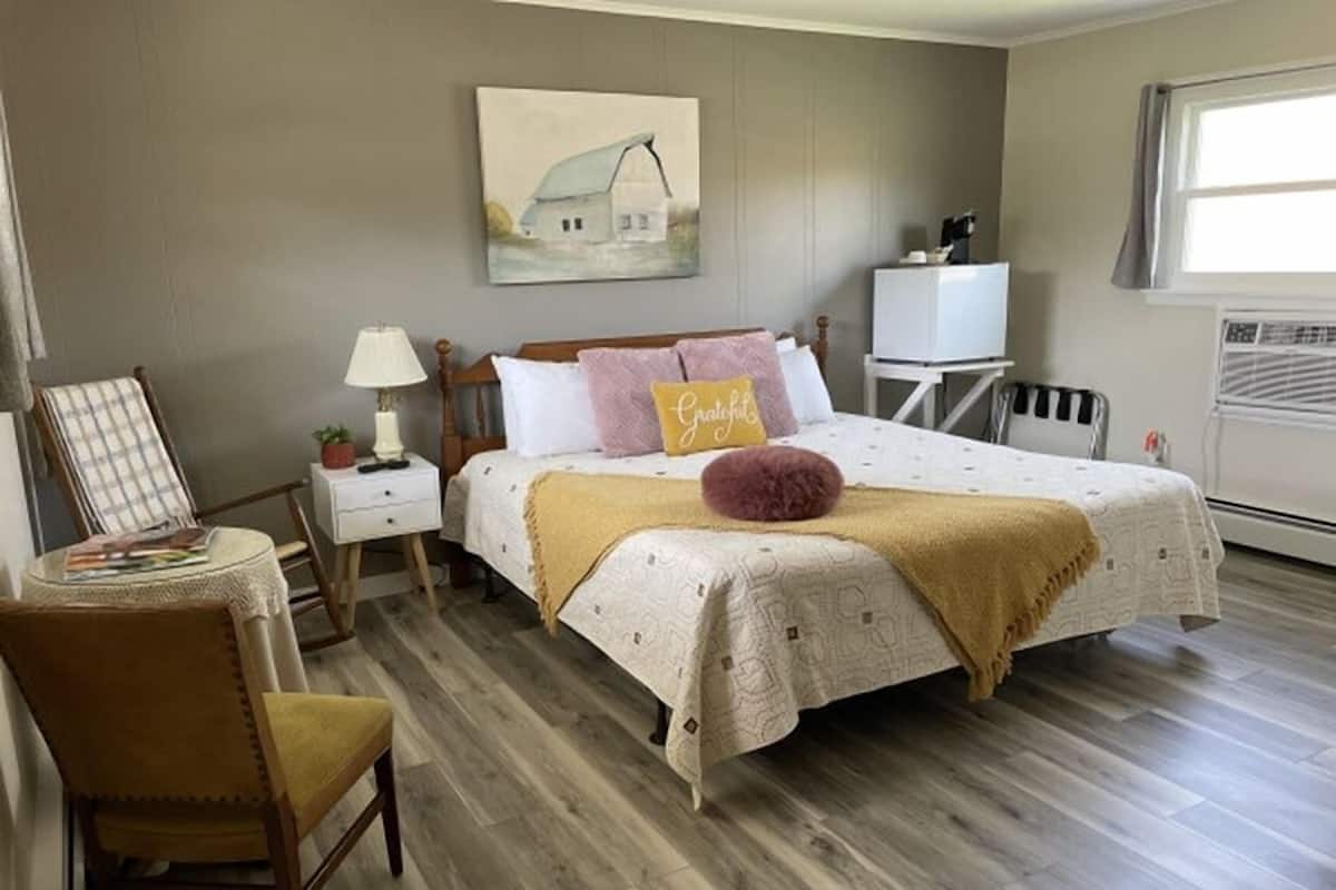 Quarto com cama, cadeira, mesa de cabeceira, luminária, quadro e janela.