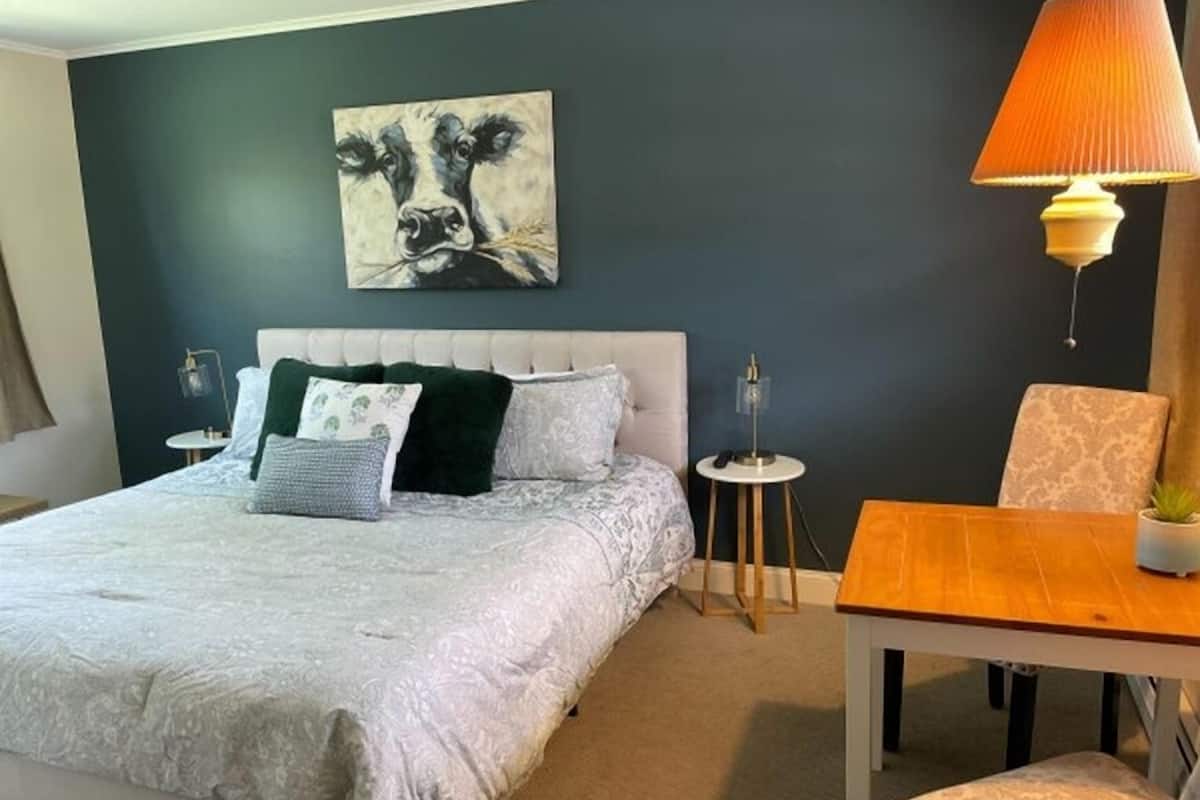 Um quarto com cama, mesa de cabeceira, abajur, cadeira e um quadro na parede.