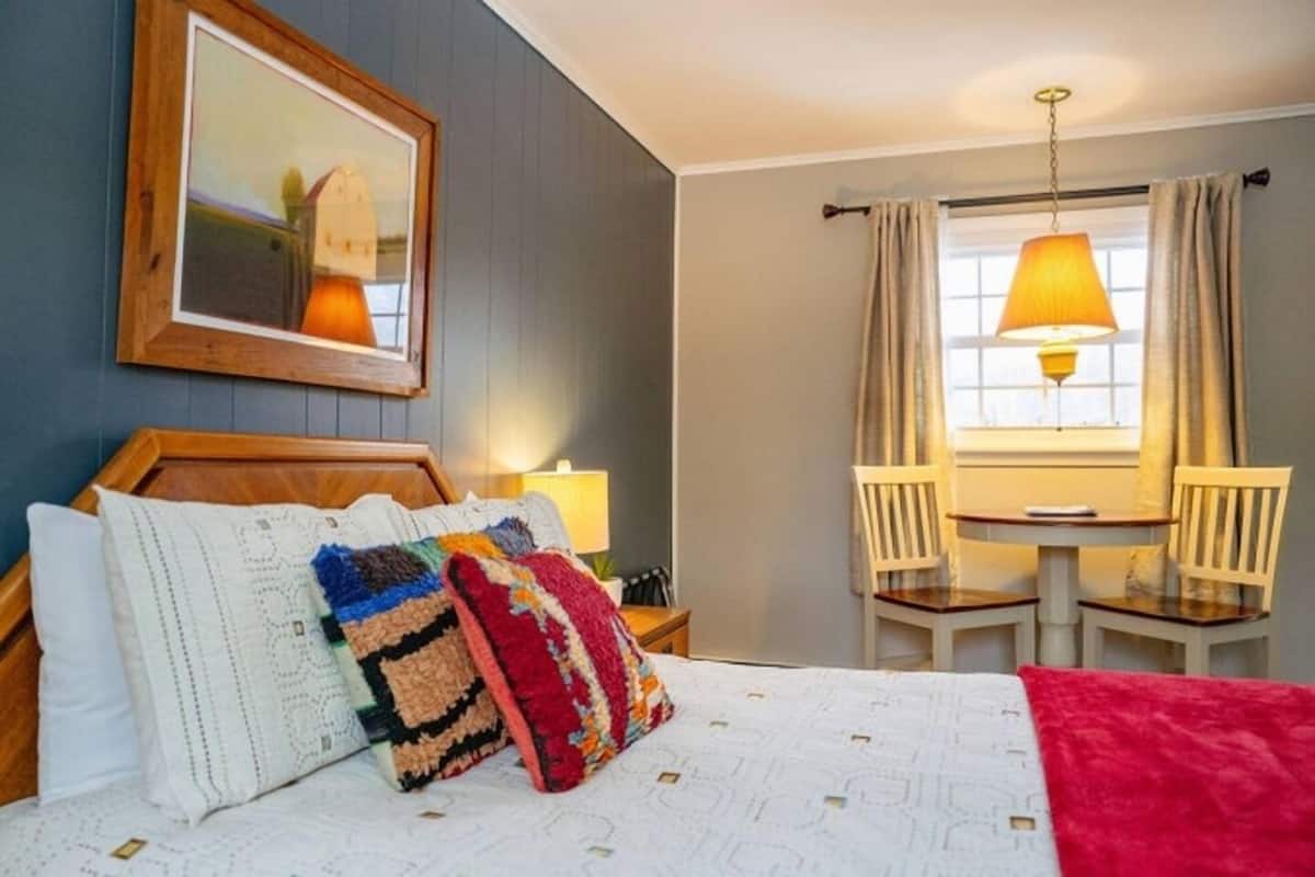 Quarto de hotel com uma cama, mesa de cabeceira com luminária, uma cadeira, uma mesinha e uma janela com cortinas.
