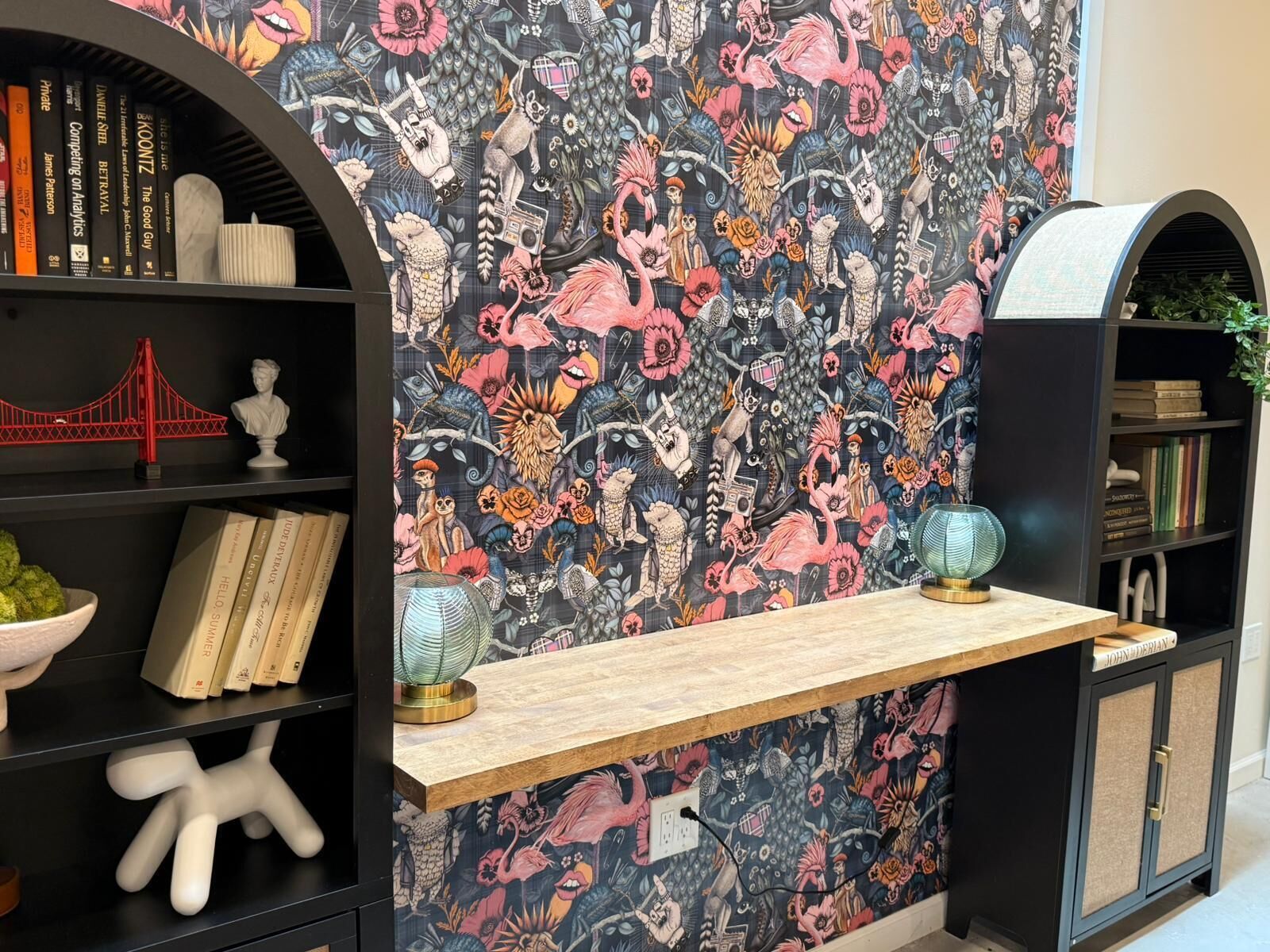 Quarto com papel de parede floral vibrante, uma prateleira de madeira com objetos decorativos e uma estante com livros e um modelo de ponte vermelha.
