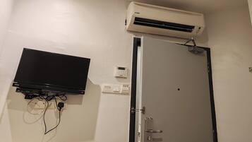 Quarto com televisão de tela plana fixada na parede, aparelho de ar condicionado acima e porta com maçaneta e tranca.