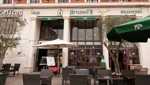 Uma área de estar ao ar livre em frente a um edifício com placas 'Brussels' e 'Bar', com mesas e guarda-sóis.