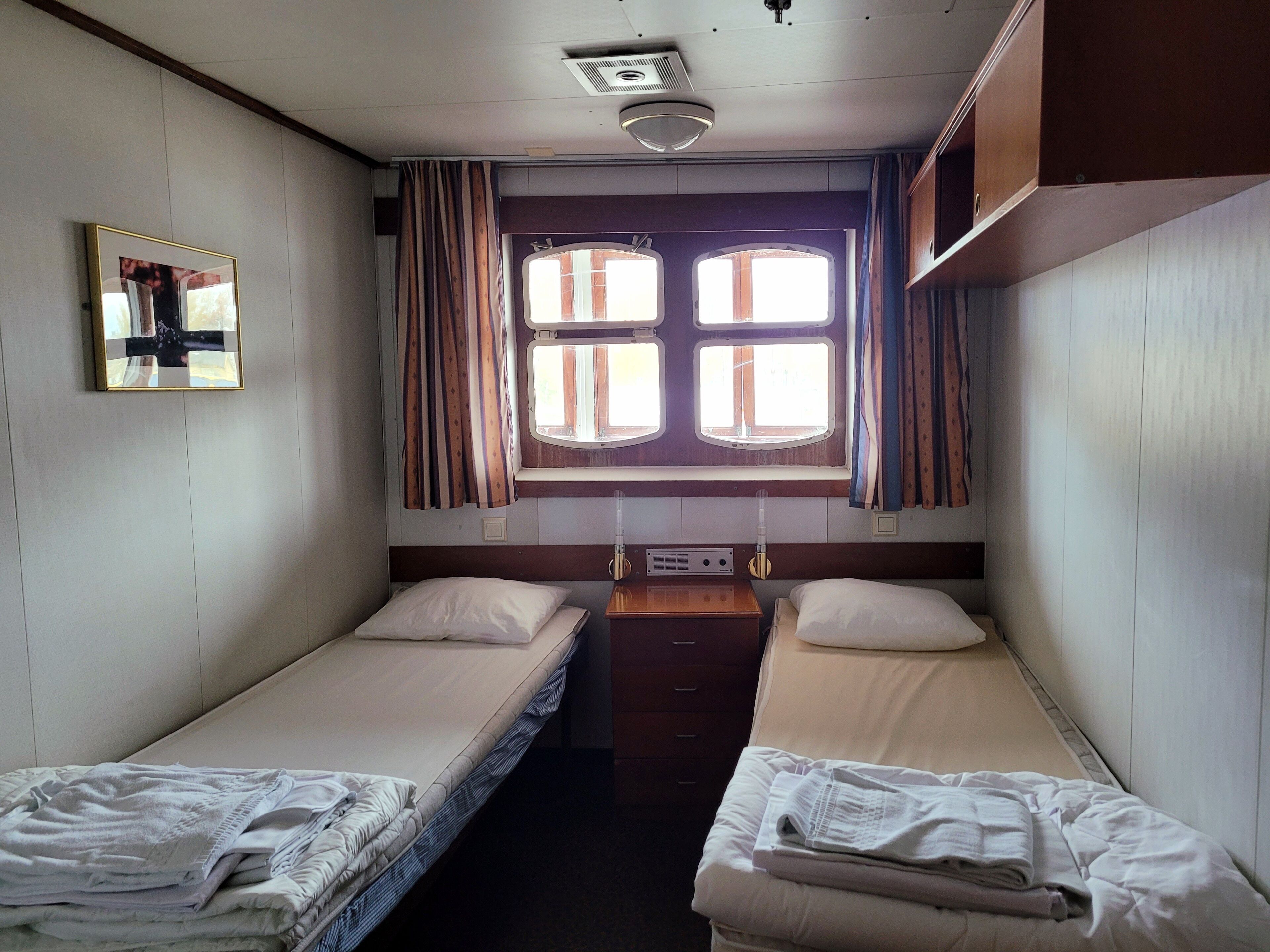Quarto pequeno e compacto com duas camas de solteiro, uma janela com cortinas, mesa de cabeceira e espelho na parede.