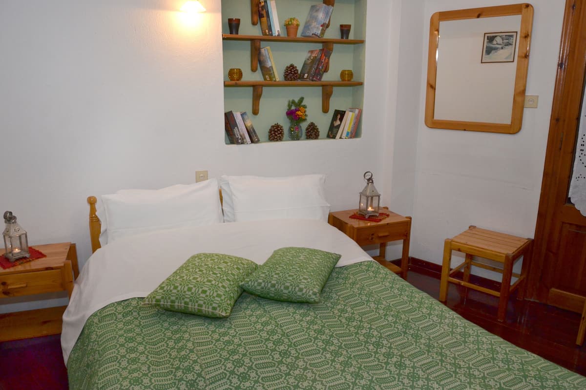 Uma cama bem arrumada com roupa de cama estampada em verde, um criado-mudo de madeira com um candeeiro e uma estante com livros e objetos decorativos.