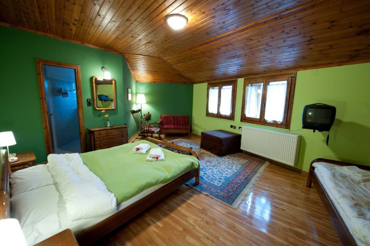Quarto com teto de madeira, cama com colcha verde, cómoda de madeira, uma cadeira, mesa pequena com luminária, radiador e televisão.