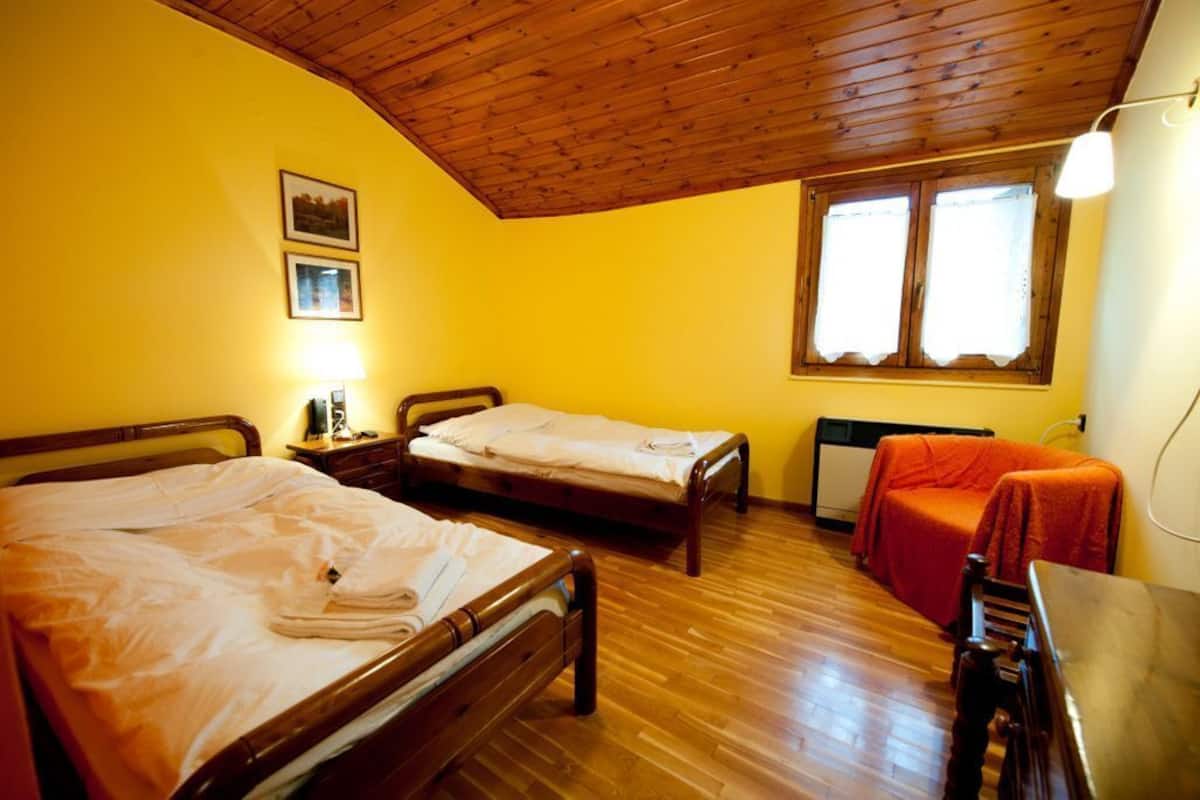 Quarto com duas camas de madeira, uma mesa pequena, uma cadeira, um radiador e uma janela com cortinas.