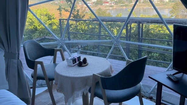 Quarto com uma janela grande, uma mesa redonda com duas cadeiras, uma cama e uma televisão.